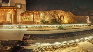 Um edifício de pedra com telhado de palha, iluminado por luzes quentes, e um caminho pavimentado que leva a uma área de piscina.