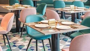 Interior de restaurante com cadeiras verde-água e rosa, mesas redondas e um chão com padrão geométrico.