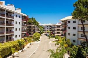 Um edifício residencial de vários andares, com varandas, área de estacionamento e vegetação ao redor.