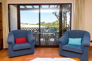 Quarto com duas poltronas, porta de vidro que dá para área externa, e vista para um prédio e árvores.