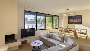 Uma sala de estar moderna com uma janela grande, uma TV de tela plana, uma lareira, um sofá e um espaço de jantar com cadeiras e uma mesa.
