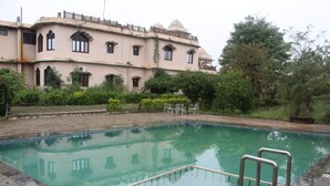 Uma piscina com escada metálica, rodeada por um jardim e um edifício com janelas em arco e uma cúpula.