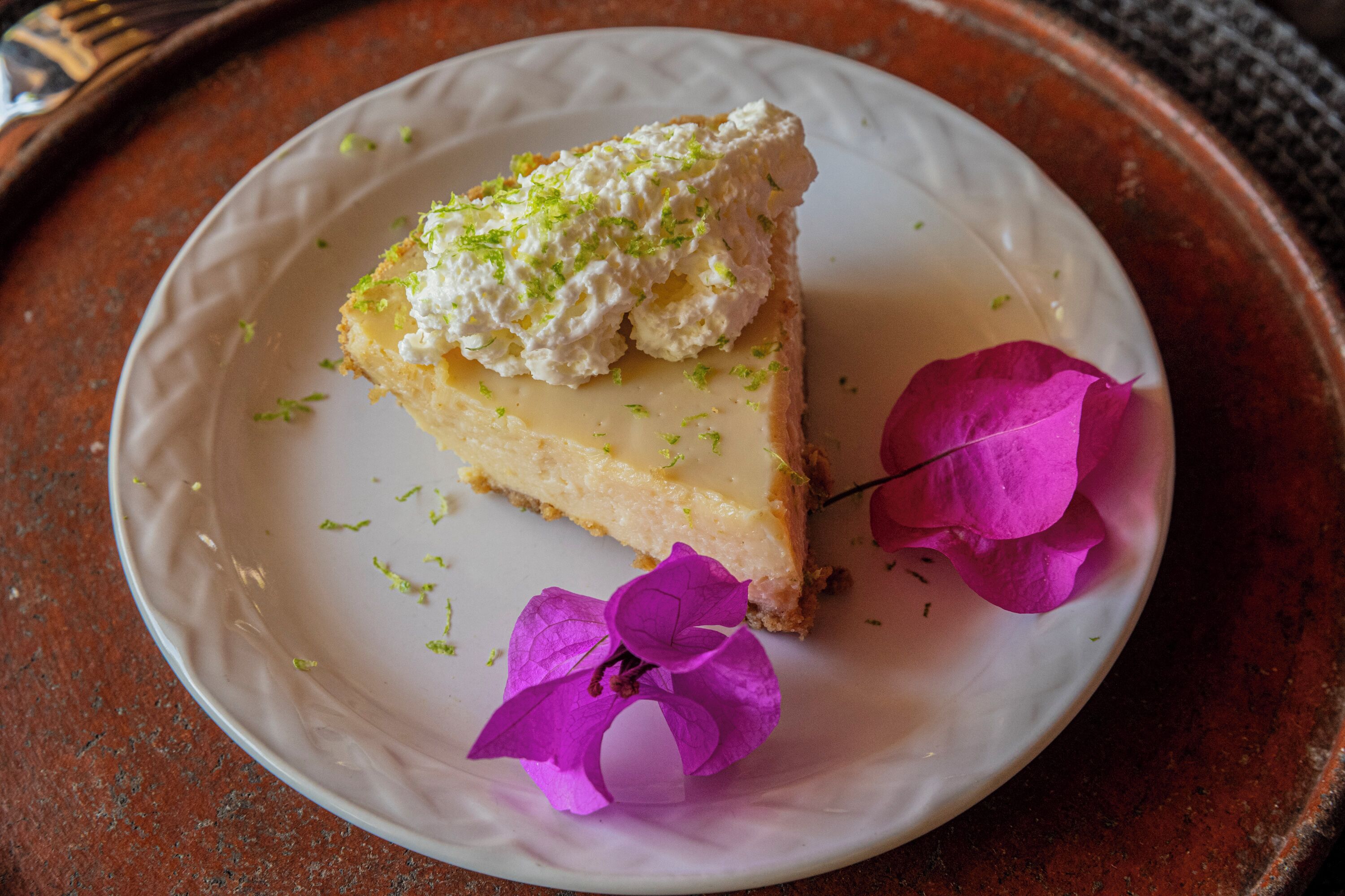 Uma fatia de cheesecake com chantilly e uma flor roxa sobre um prato.