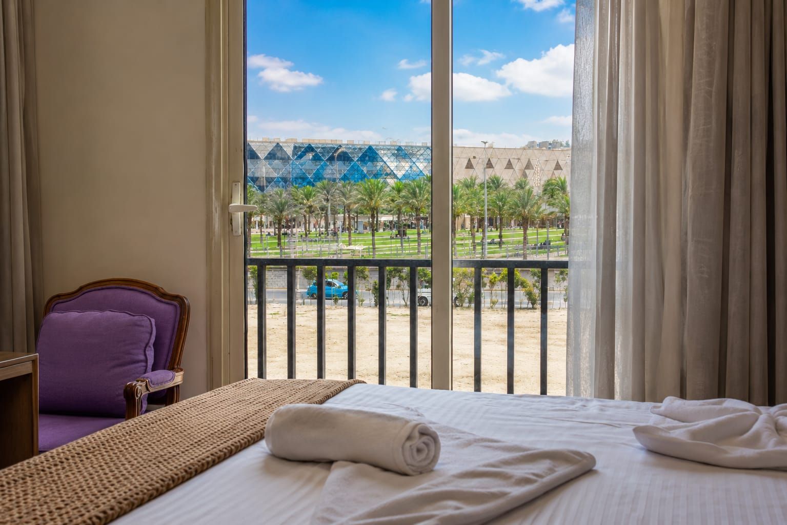 Quarto de hotel com cama, uma poltrona roxa e vista para um edifício azul e palmeiras pela janela.