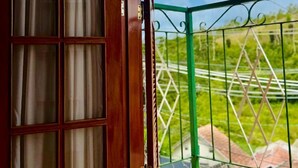 Uma porta aberta dá para uma varanda com vista para uma paisagem verde e um céu nublado.