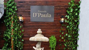 Uma fonte de pedra com uma estátua, rodeada por vegetação, com um muro de madeira e uma placa com a inscrição 'Pousada D'Paula'.