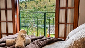 Uma cama com um cobertor marrom e um travesseiro branco, um ursinho de pelúcia e vista para uma colina verde através de uma varanda com grade de metal.