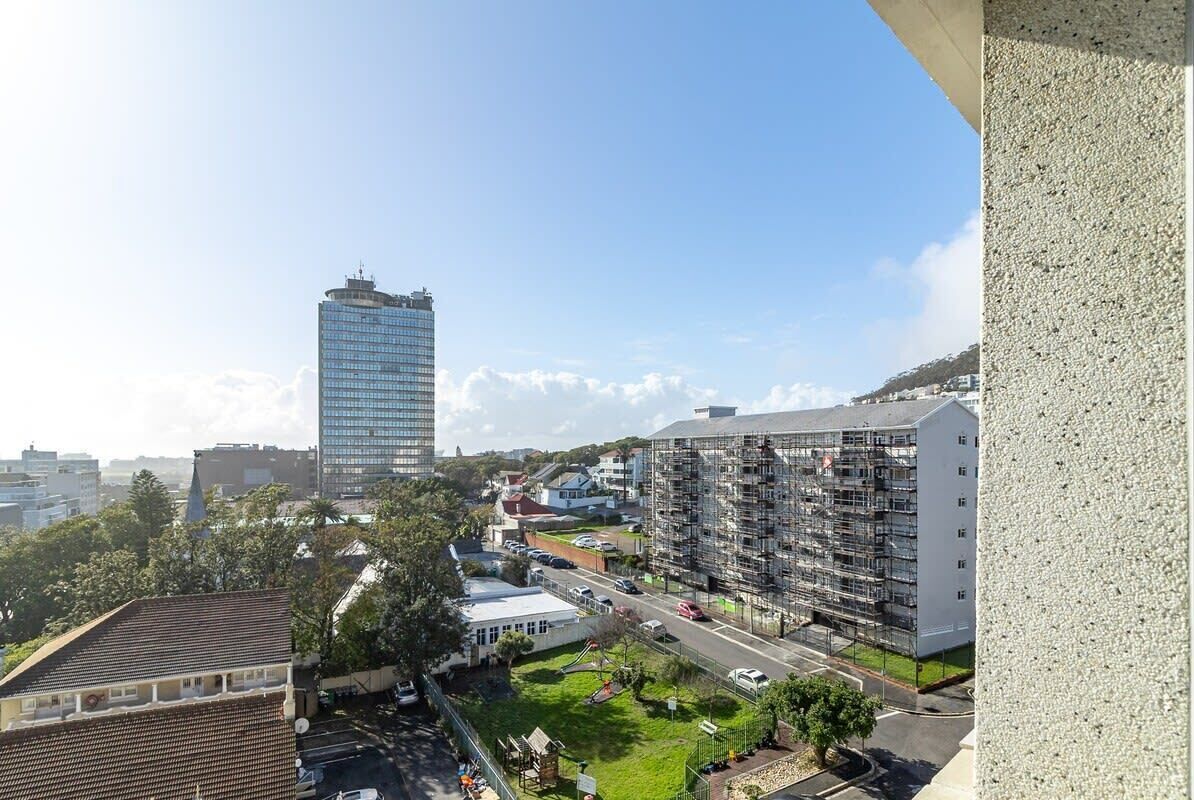 Um edifício alto com fachada de vidro moderna, cercado por edifícios residenciais e uma área verde de parque.