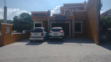 Entrada de hotel com dois carros estacionados na frente, uma placa com a inscrição "HOTEL EL REFUGIO" e uma placa menor para "PRINCON MAGICO".