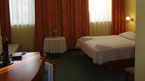 Quarto de hotel com uma cama, uma escrivaninha com computador, uma luminária e uma pequena mesa com um vaso.