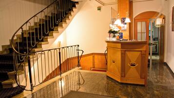 Um saguão de hotel com uma escadaria de mármore, um balcão de recepção e uma área de atendimento em madeira.