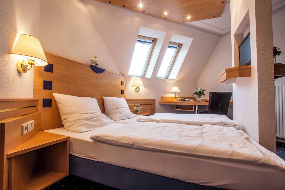 Quarto de hotel com duas camas, uma escrivaninha, uma cadeira e duas janelas.