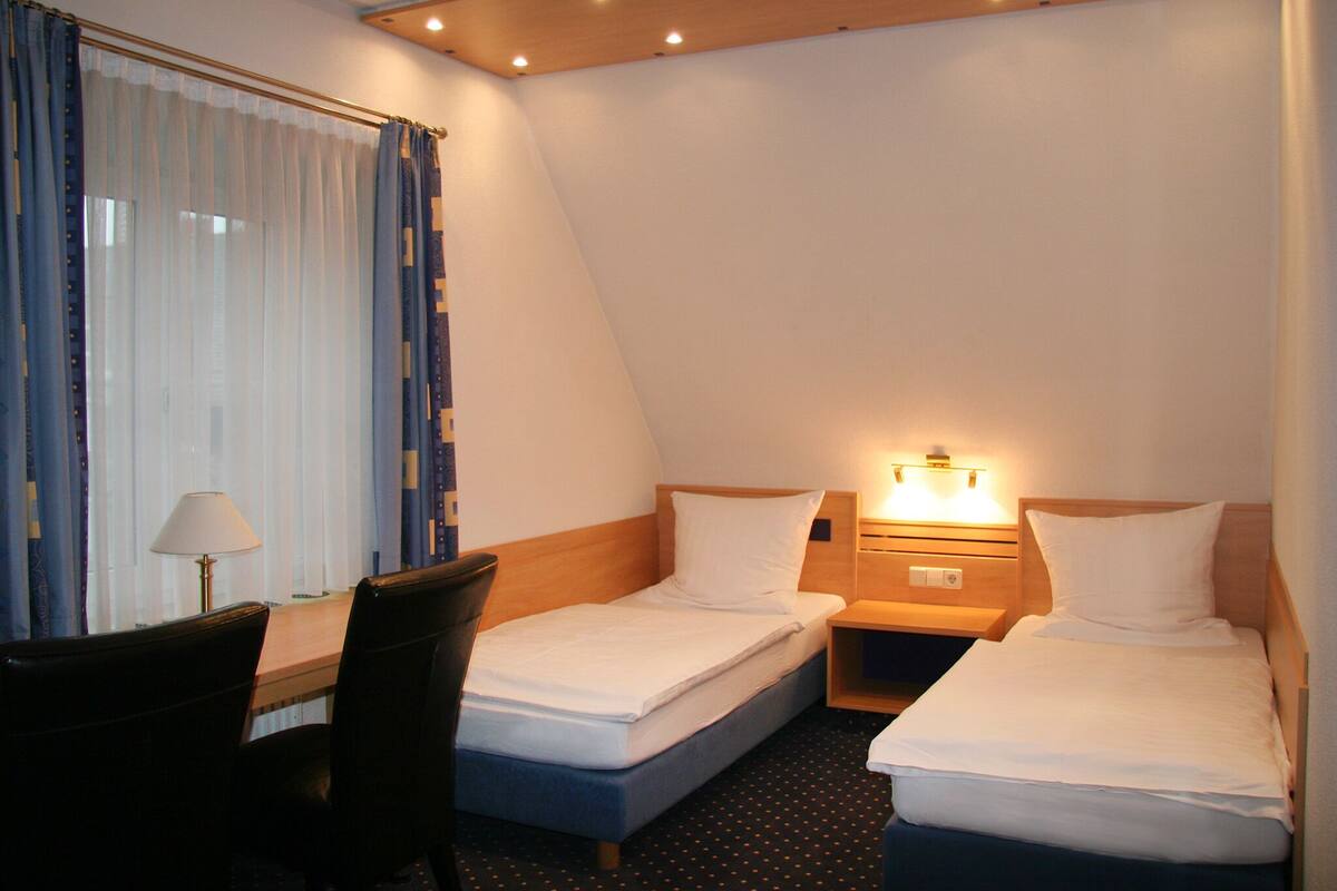 Quarto de hotel com duas camas de solteiro, uma escrivaninha com cadeira e uma janela com cortinas.