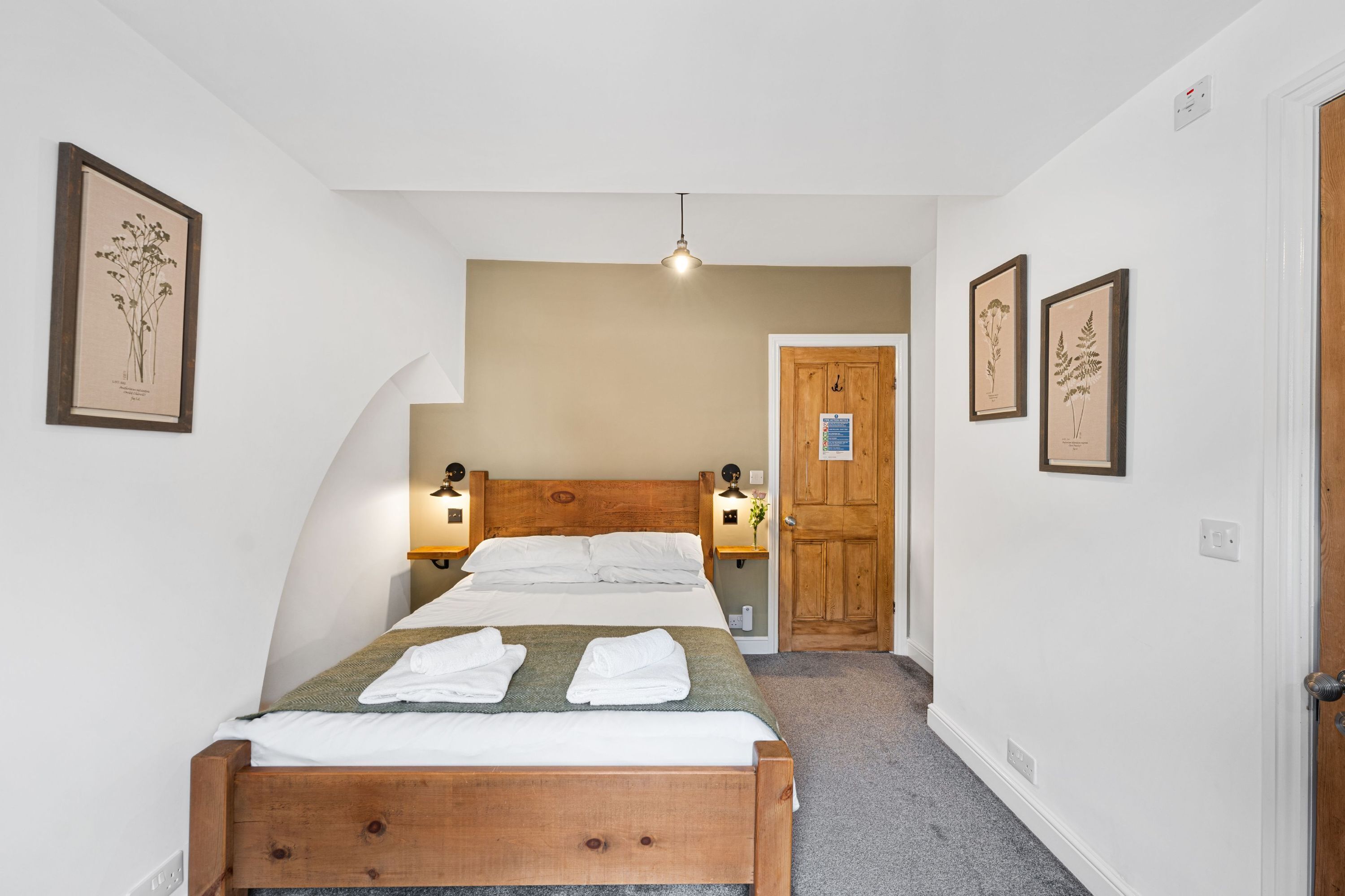 Um quarto com uma cama de madeira, duas mesas de cabeceira com abajures, uma porta e gravuras botânicas emolduradas nas paredes.