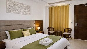 Quarto de hotel com cama, mesa de cabeceira, abajur, duas cadeiras, uma mesa e uma porta.
