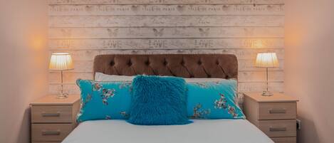 Quarto com cabeceira de madeira, duas mesas de cabeceira com luminárias, cama com roupa de cama florida e um travesseiro azul macio.
