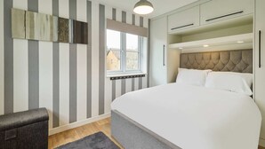 Quarto moderno com cama grande, um banquinho estofado cinza, tapete felpudo cinza e parede listrada com janela.