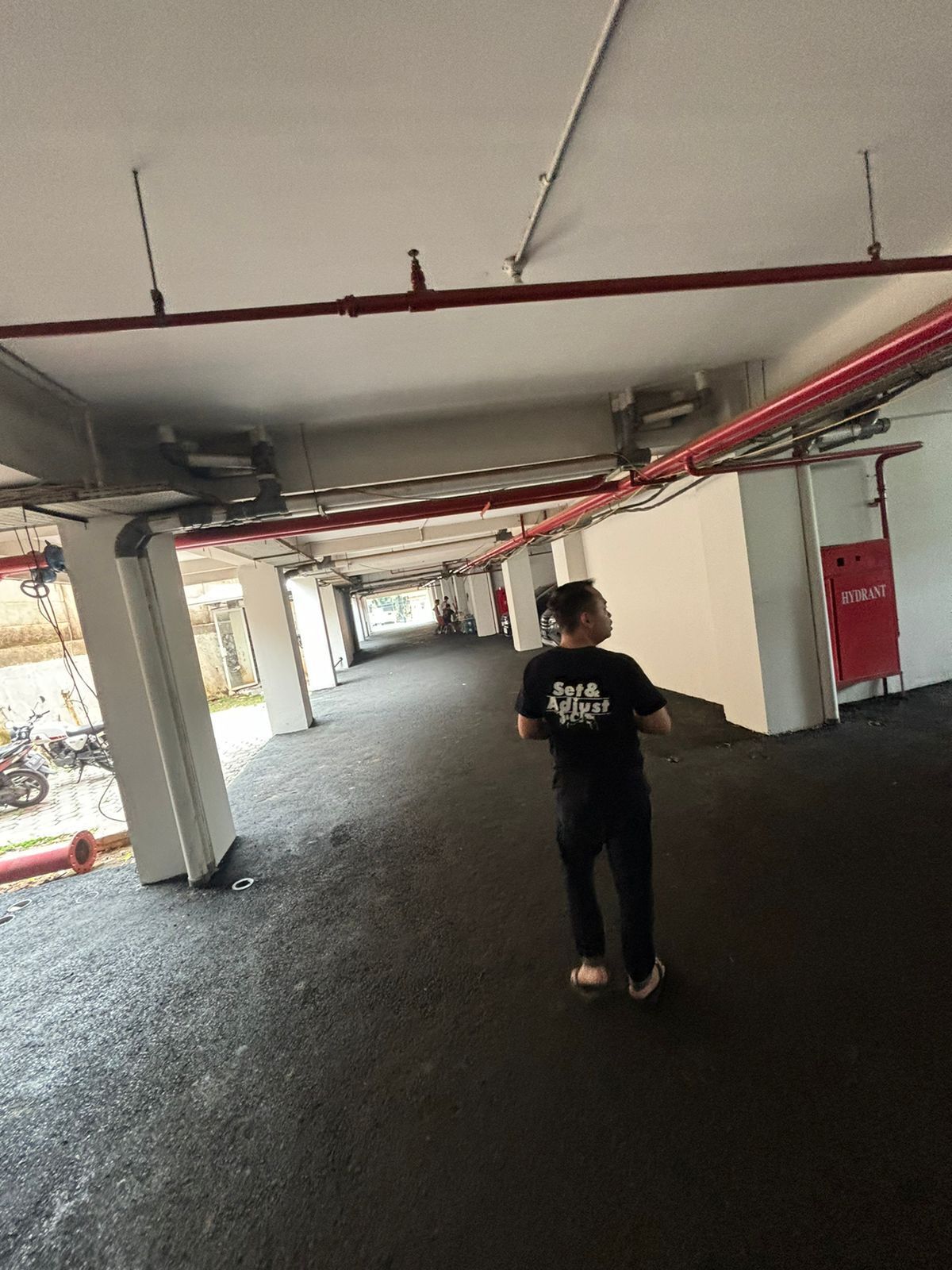 Uma pessoa parada numa área de estacionamento coberta, com pilares de concreto e tubos vermelhos no teto.
