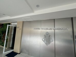 Entrada de um edifício moderno com fachada metálica e um logotipo que diz 'KOSTEL RESIDENCE GENDEKIA'.