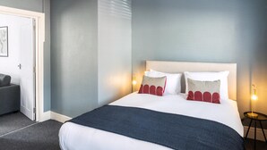 Cama bem arrumada com cabeceira branca, dois travesseiros e um cobertor escuro. Há uma mesa de cabeceira com abajur, uma porta com moldura de quadro e um sofá cinza ao fundo.