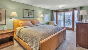 Quarto com uma cama grande, duas luminárias de cabeceira, mesa de cabeceira, cômoda de madeira e porta de vidro corrediça que dá para um pátio externo.