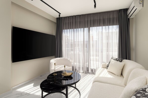 Uma sala de estar moderna com uma TV de tela grande, um sofá, uma mesa de centro e cortinas leves.