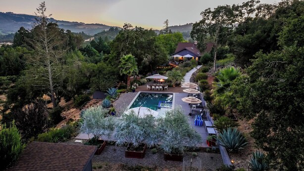 Piscina com espreguiçadeiras e guarda-sóis ao redor, em meio a jardins e árvores ajardinadas.