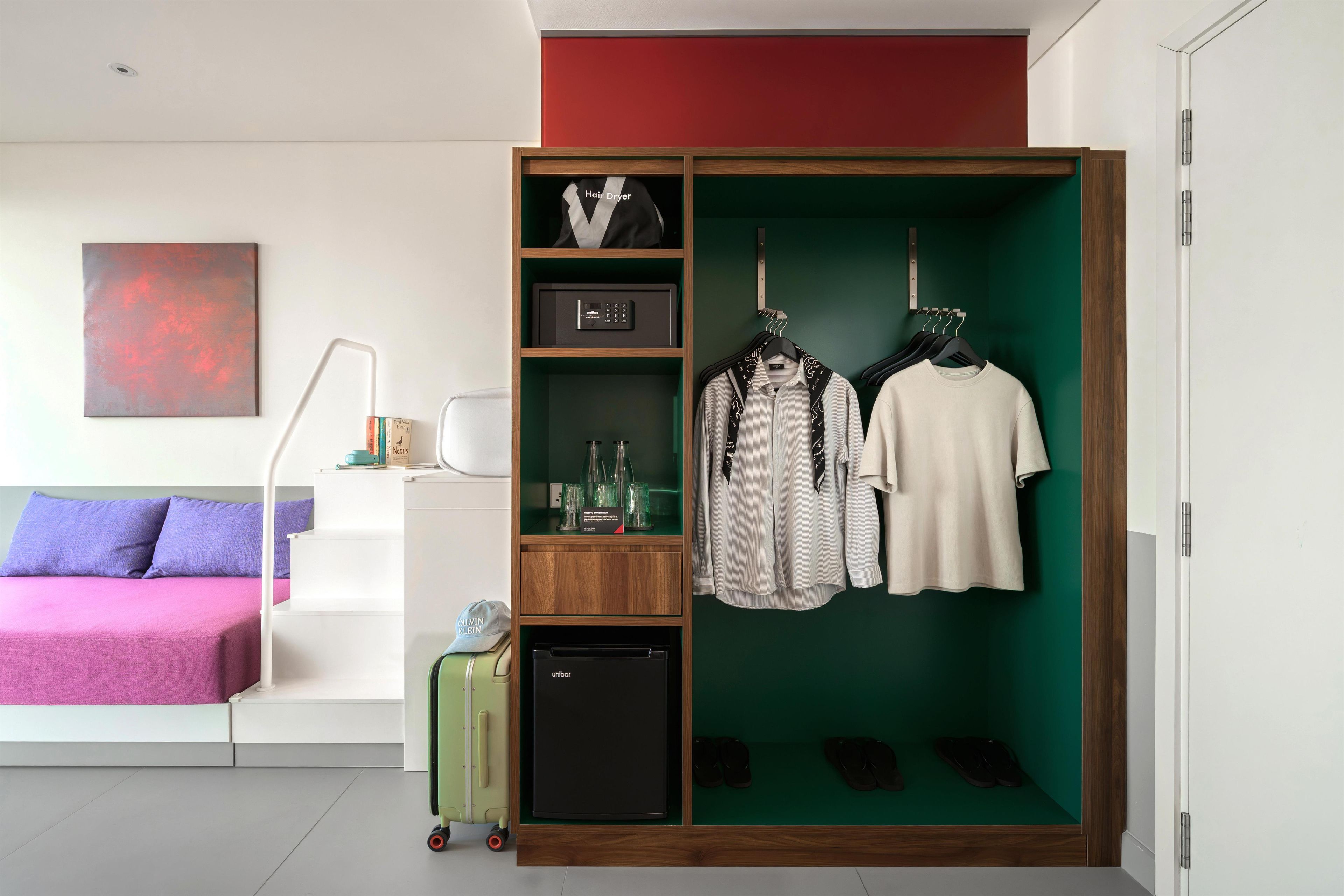 Quarto de hotel com cama, escrivaninha, guarda-roupa e mala.