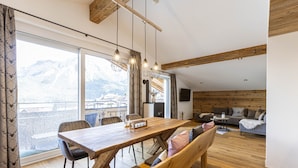 Uma ampla área de jantar com mesa e bancos de madeira, janelas grandes com vista para as montanhas e uma área de estar aconchegante com sofá e lareira.