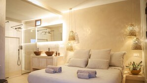 Quarto bem iluminado com cama grande, luminárias de cabeceira, espelho e banheiro com chuveiro e pia.