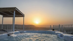 Uma banheira de hidromassagem com um deck de madeira e guarda-corpos de metal, com vista para o pôr do sol sobre a água.
