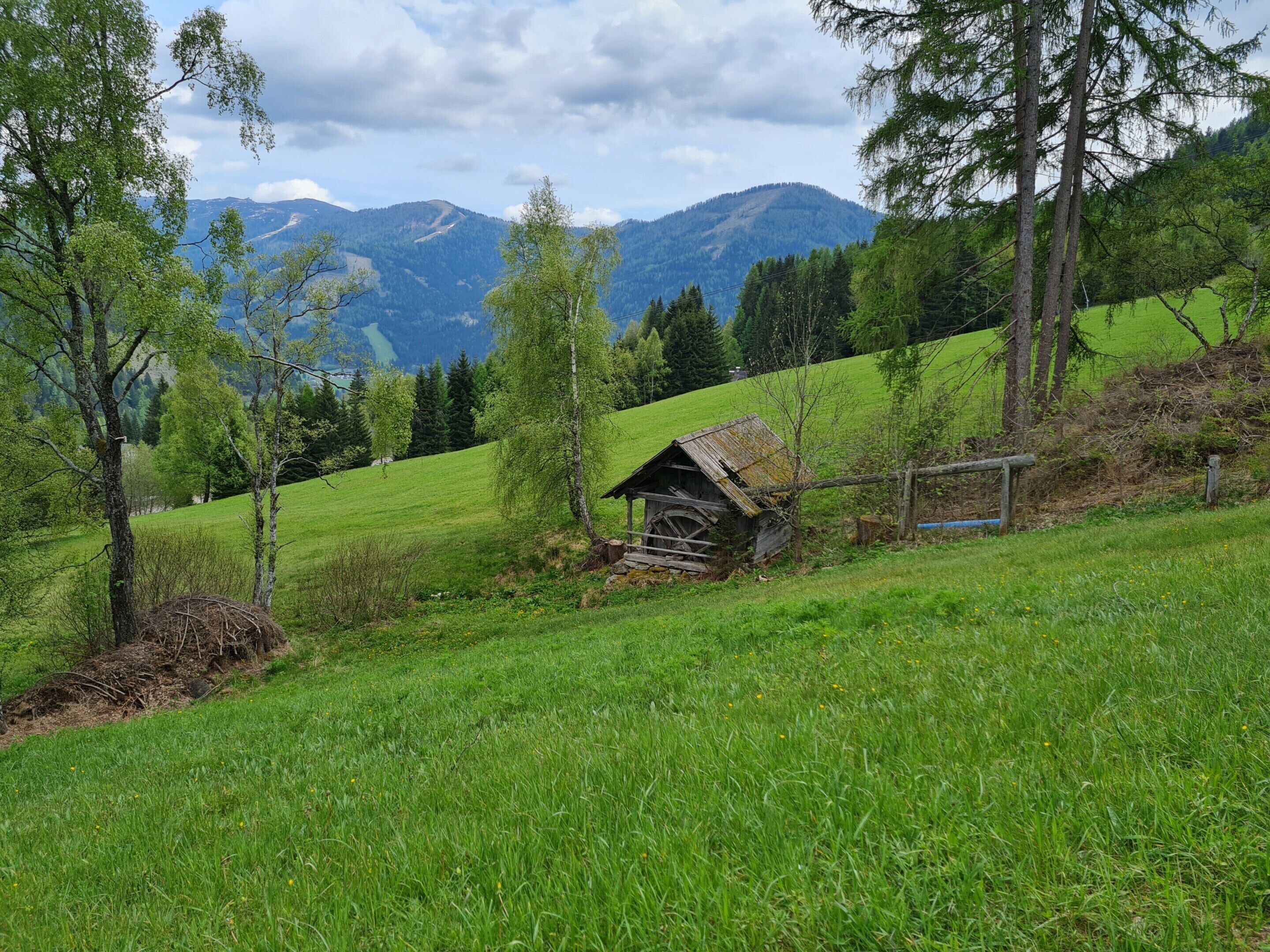 Uma cabana rústica de madeira num campo verdejante, com montanhas ao fundo.
