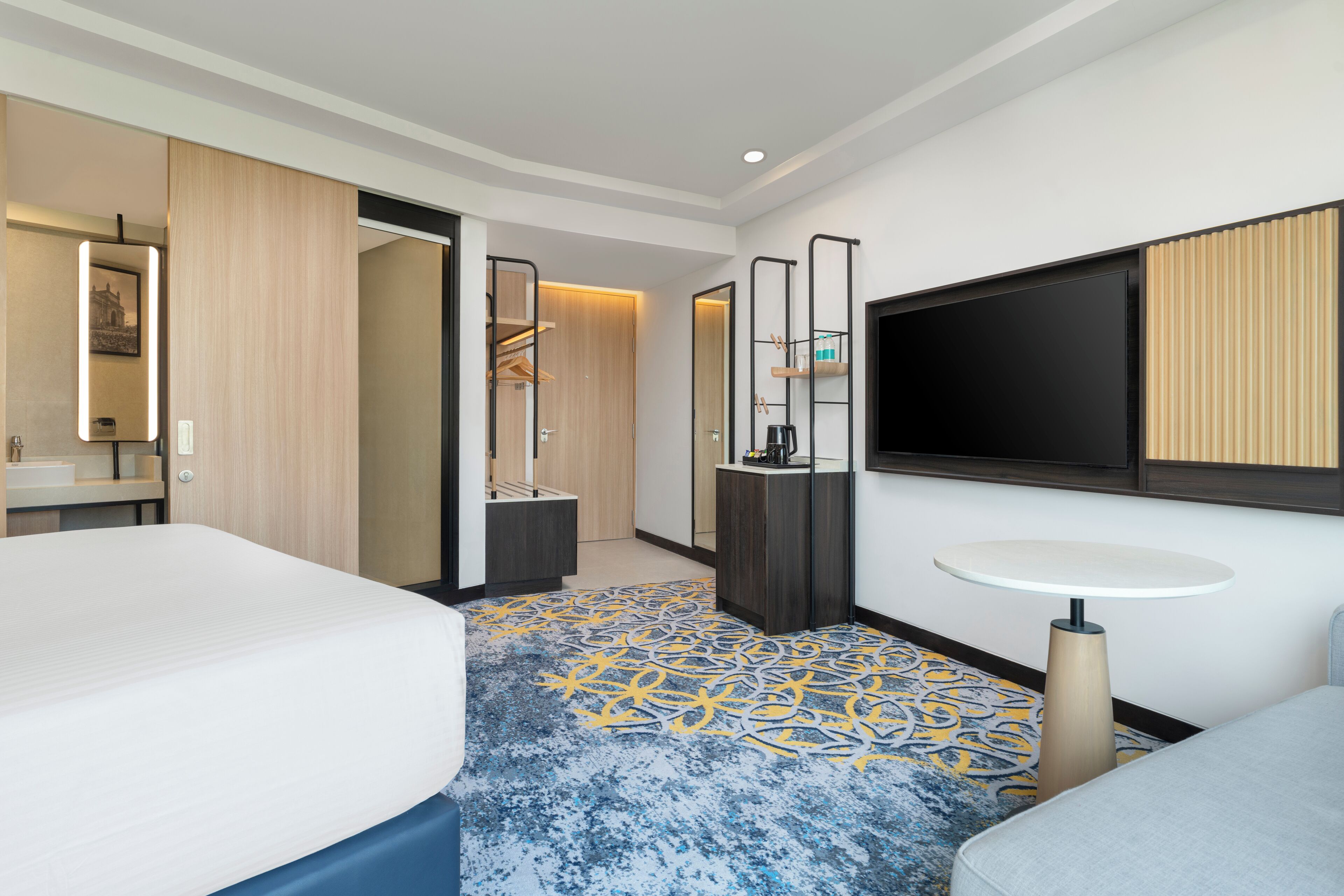 Quarto de hotel com cama, escrivaninha, espelho, televisão e banheiro.