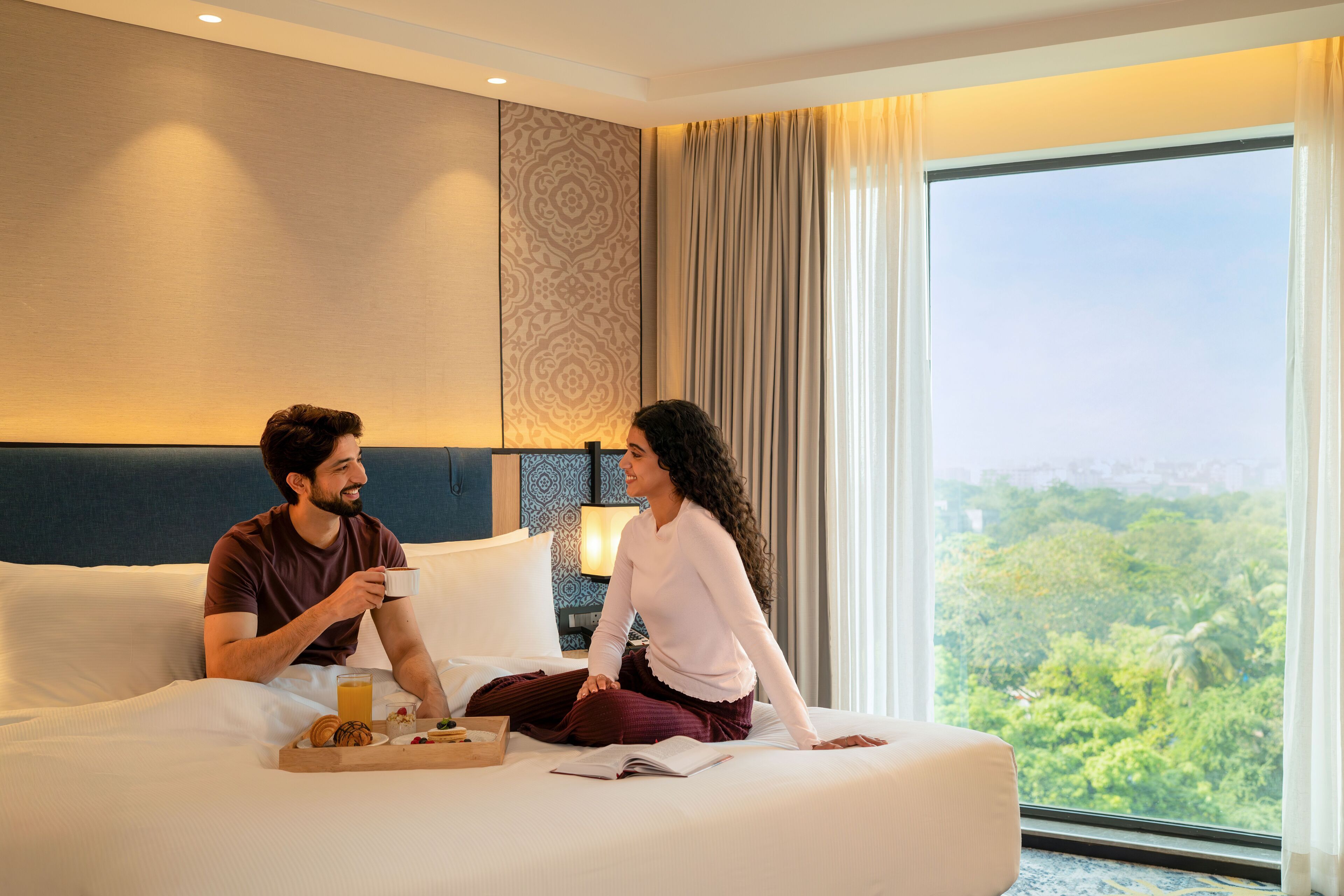 Um casal sentado em uma cama em quarto de hotel com vista para a cidade, uma bandeja de café da manhã e uma mesa de cabeceira com abajur.