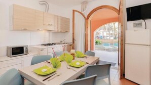 Cozinha compacta com micro-ondas, fogão e armários. Uma mesa de jantar está posta com jogos americanos e talheres verdes. Uma porta aberta mostra uma área de piscina com cadeiras.