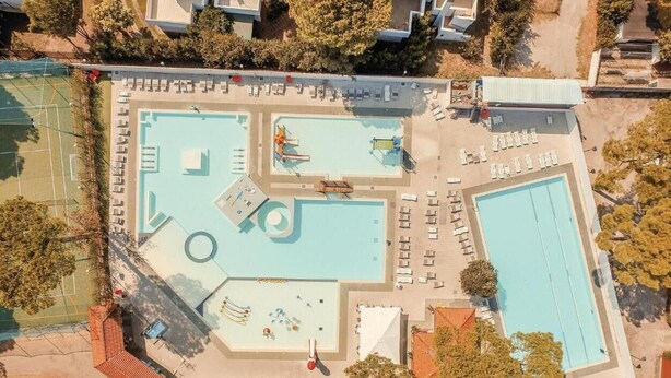Vista aérea de um complexo de piscinas com várias piscinas, espreguiçadeiras e uma quadra de tênis.