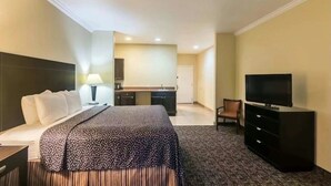 Quarto de hotel com uma cama, televisão, escrivaninha com luminária e uma kitchenette ao fundo.