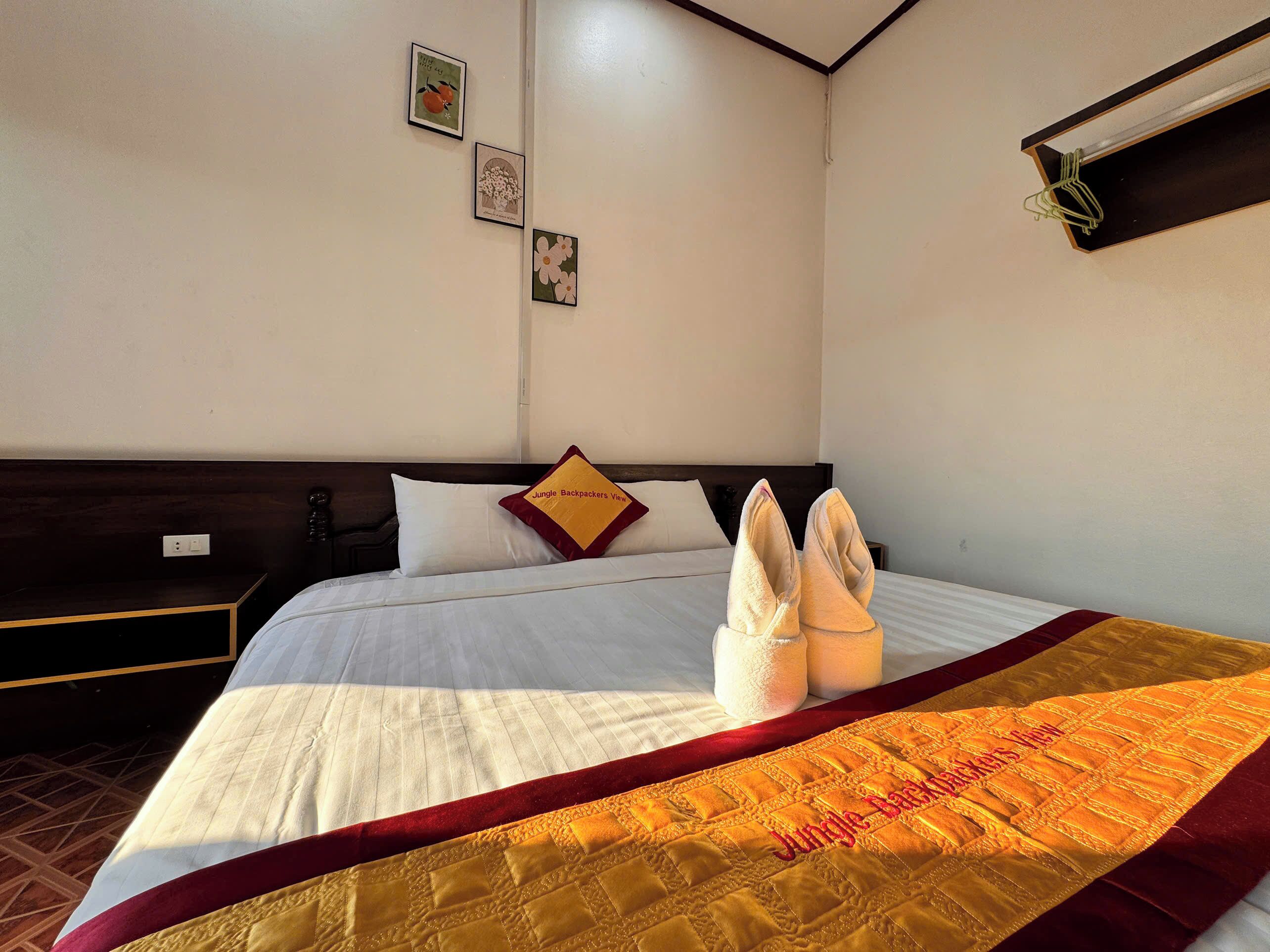 Quarto de hotel com cama bem arrumada, manta vermelha e dourada, e almofada com a inscrição 'Jungle Backpackers View'.