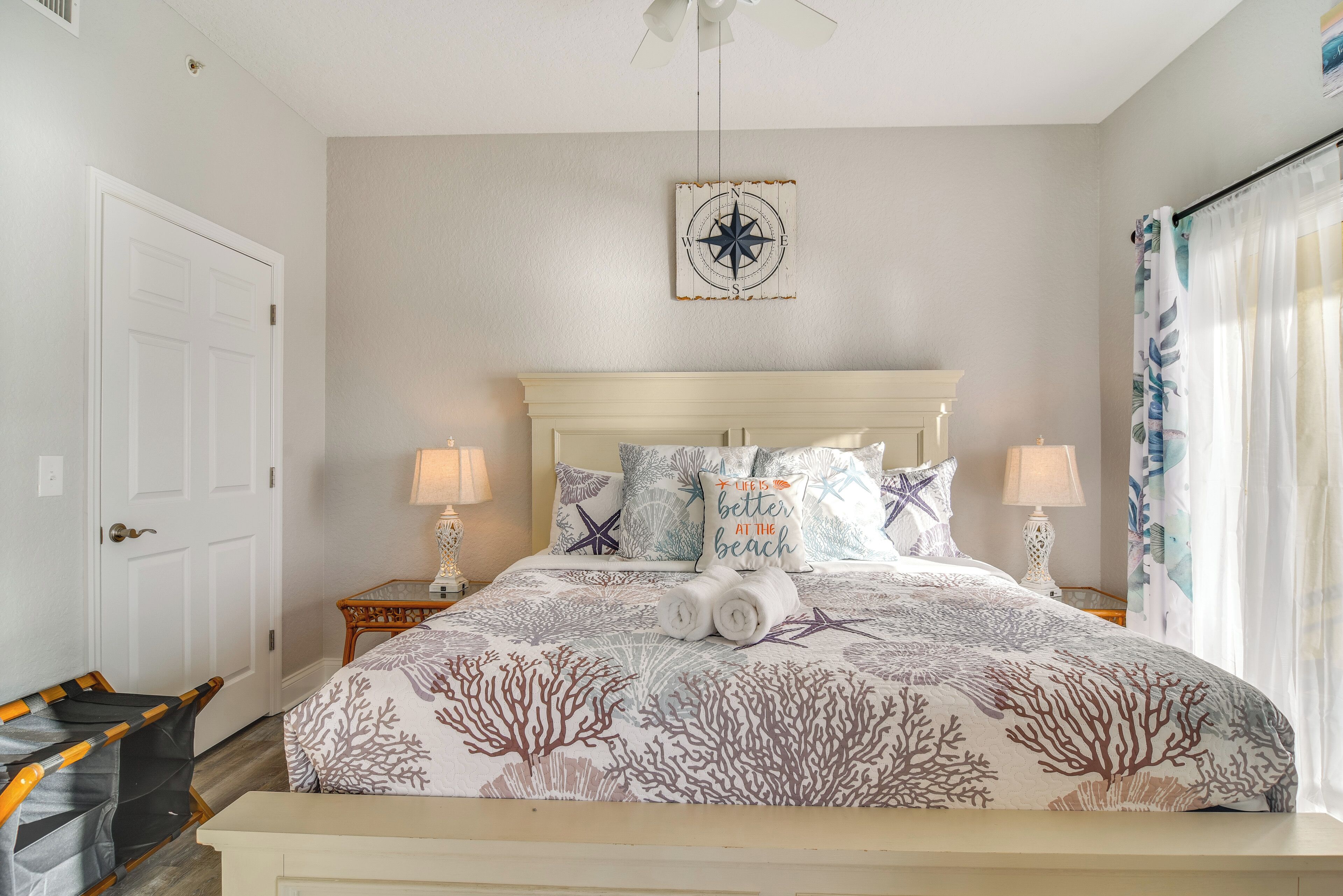 Quarto com cama, mesa de cabeceira, luminária, ventilador de teto e decoração de parede com bússola.