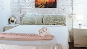 Quarto com parede de tijolos brancos, uma pintura, cama com toalha rosa e mesa de cabeceira com luminária.