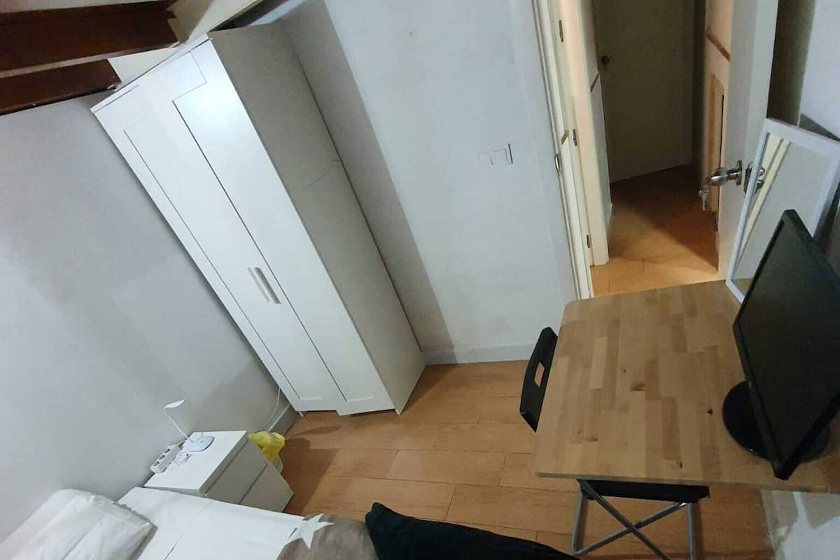 Quarto compacto com uma cama, uma pequena escrivaninha, uma cadeira e uma televisão. Há uma porta que dá para outro cômodo.