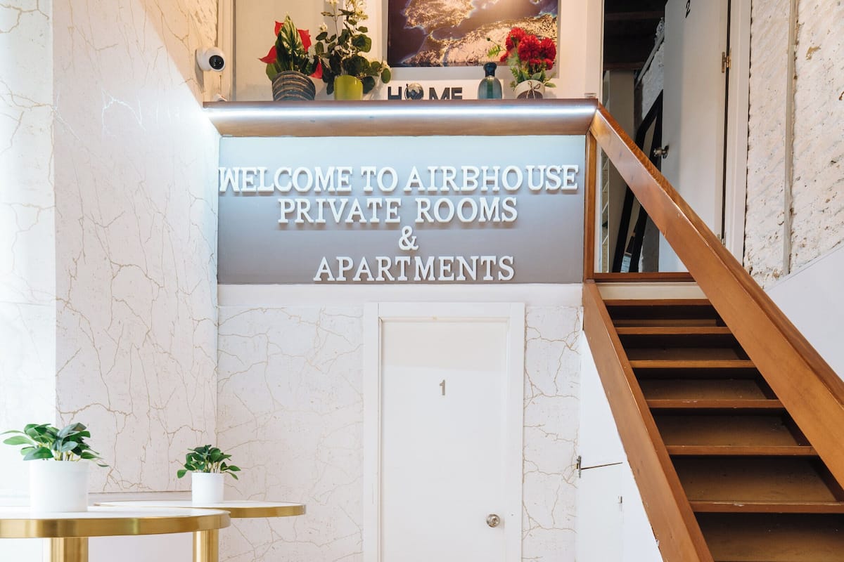 Um interior moderno com uma placa exibindo 'Welcome to Airhouse Private Rooms & Apartments', banquetes de madeira, uma escada e uma porta com um número.
