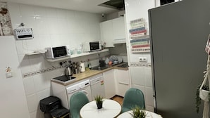 Uma cozinha compacta com armários brancos, micro-ondas, pia e geladeira. Há duas cadeiras verde-água ao redor de uma pequena mesa branca com um vaso de planta.