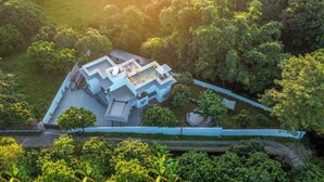 Vista aérea de uma casa moderna rodeada por vegetação densa, com céu claro e luz solar projetando sombras.
