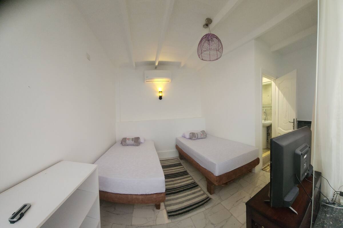 Quarto pequeno e limpo com duas camas de solteiro, televisão e banheiro visível por uma porta aberta.