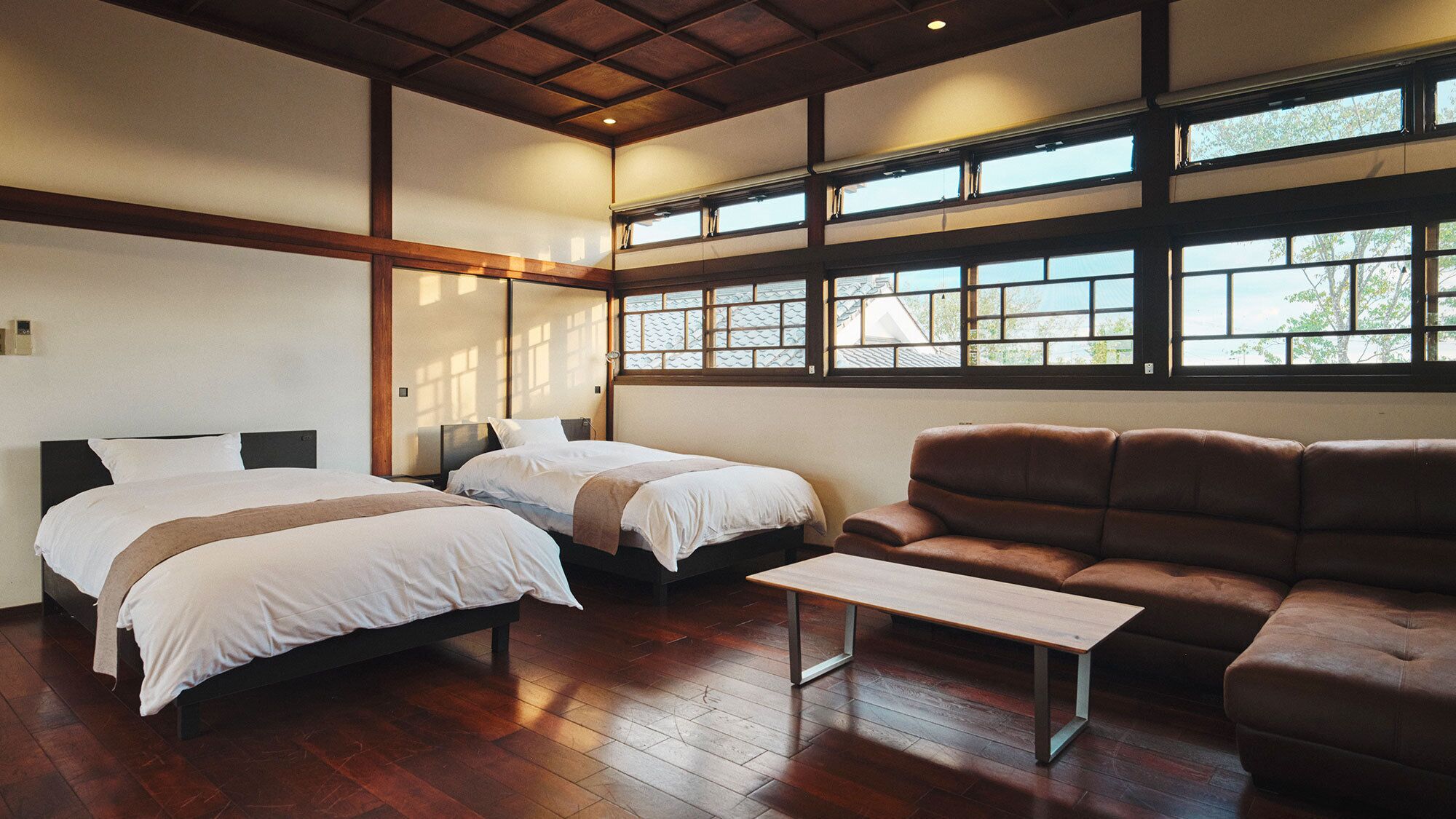Quarto de hotel com duas camas, um sofá, uma mesa pequena e janelas amplas.