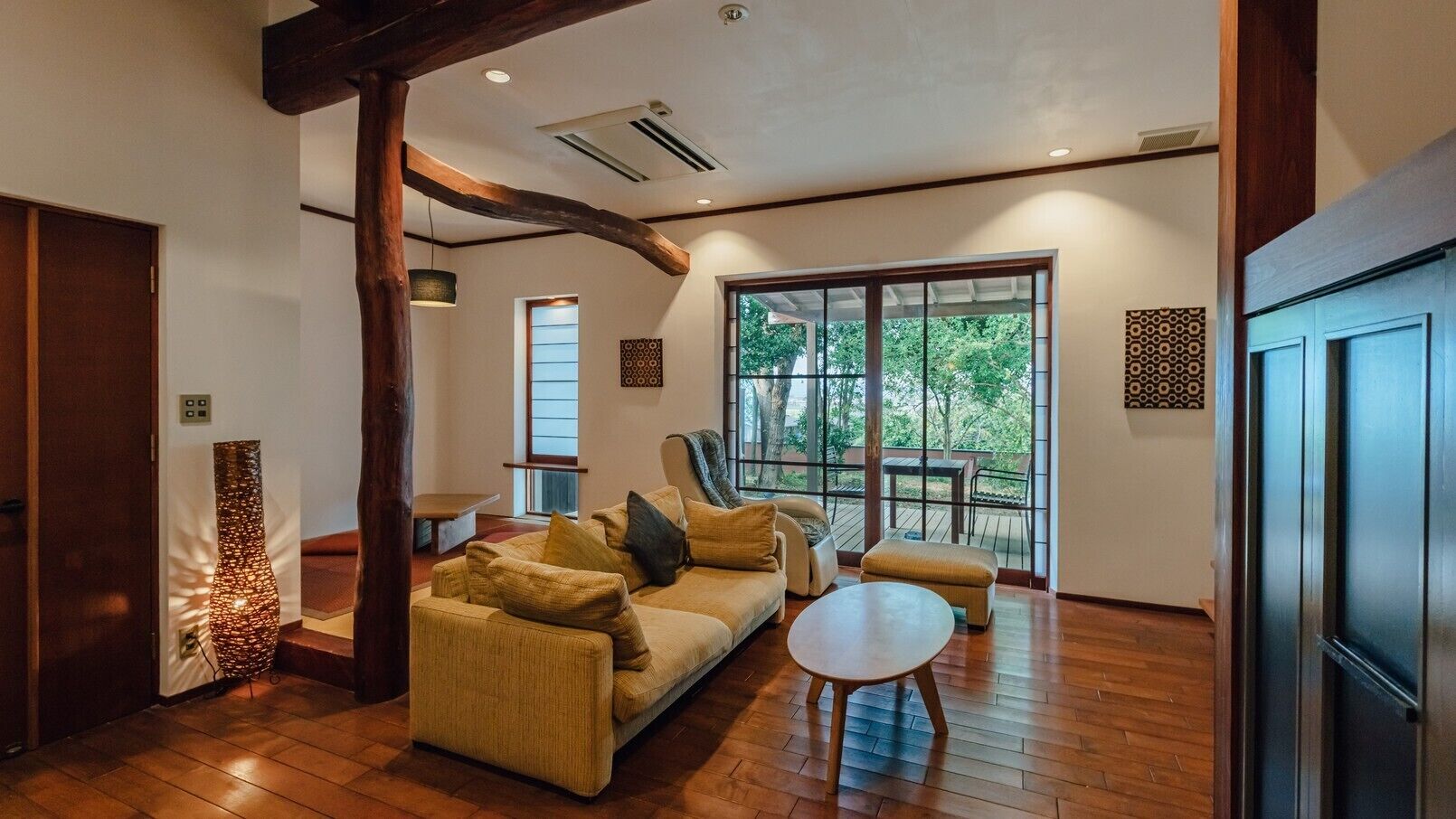 Ampla sala de estar com sofá, banqueta e mesa de centro redonda. Há uma grande janela com vista para o exterior.