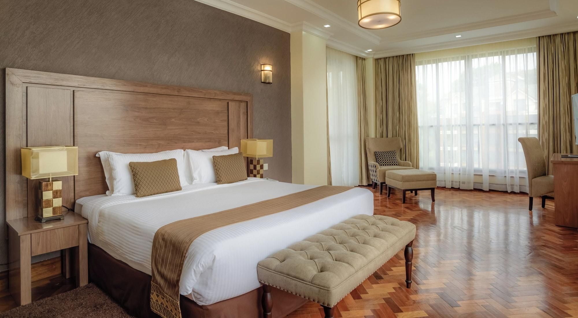 Quarto de hotel com uma cama grande, duas luminárias de cabeceira, um painel de madeira na cabeceira, um banco, uma cadeira e uma escrivaninha.