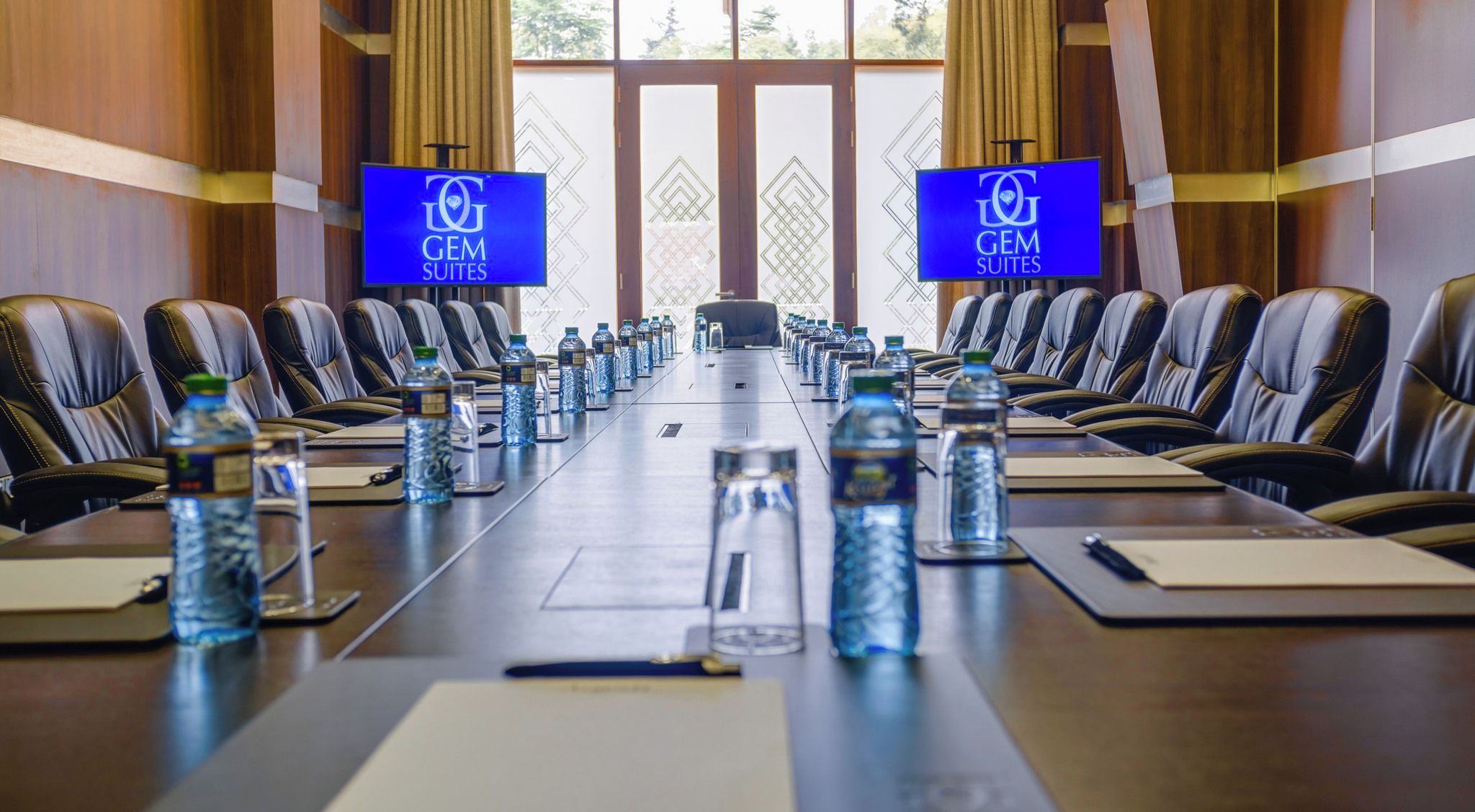 Uma sala de conferências com uma mesa comprida, várias cadeiras, garrafas de água e duas telas exibindo o logotipo 'GEM SUITES'.