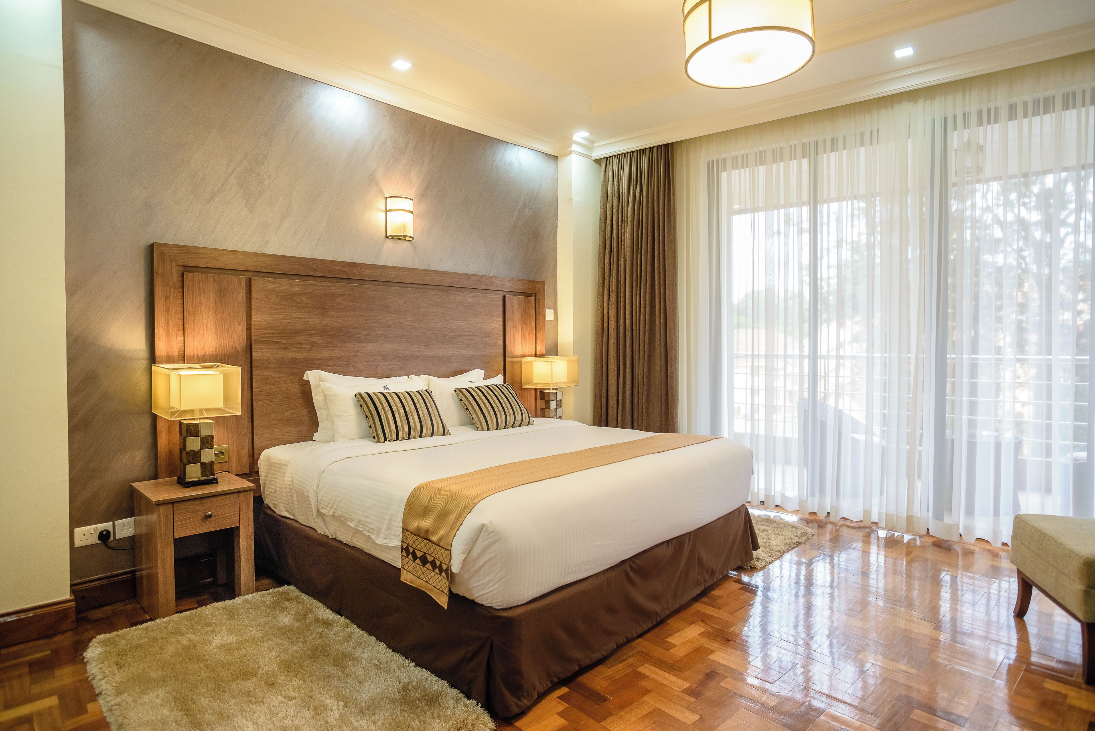 Quarto de hotel com uma cama grande, cabeceira de madeira, abajures e vista do exterior através de cortinas finas.